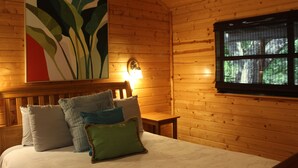 Quarto de cabana de madeira com cama, almofadas, um relógio na parede e uma janela com vista para árvores.