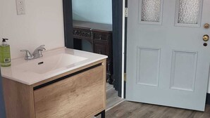 Casa de banho com toucador de madeira, lavatório branco e espelho. Tem uma porta com painéis de vidro jateado e fechadura.