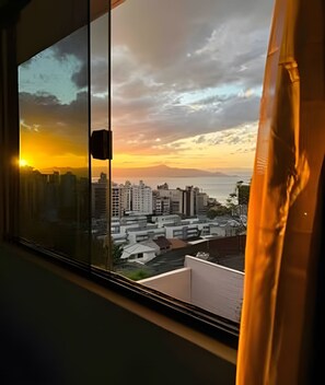 Vista pela janela, com cortina, mostrando um horizonte urbano com edifícios e um pôr do sol.