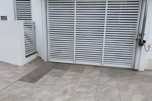 Um portão branco de metal fechado, com ripas horizontais, um chão de azulejos e uma parede branca com uma tomada.
