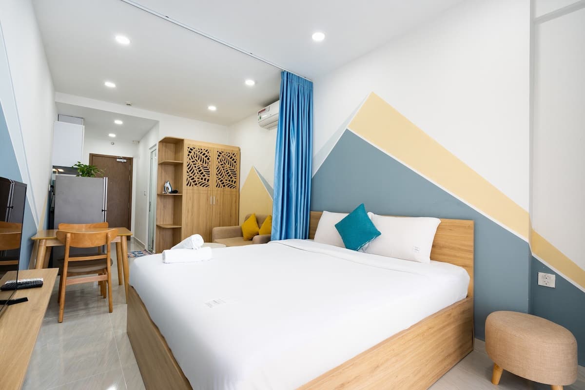 Quarto de hotel moderno com cama de madeira, uma escrivaninha com cadeira, um guarda-roupa e um banheiro visível através de uma porta aberta.