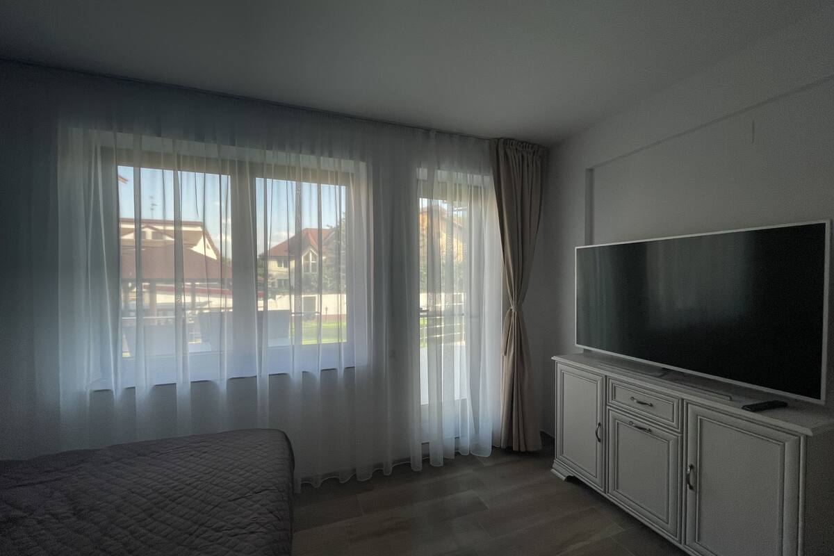 Quarto de hotel com uma grande janela, uma cama, uma televisão em um móvel e vista para as casas lá fora.