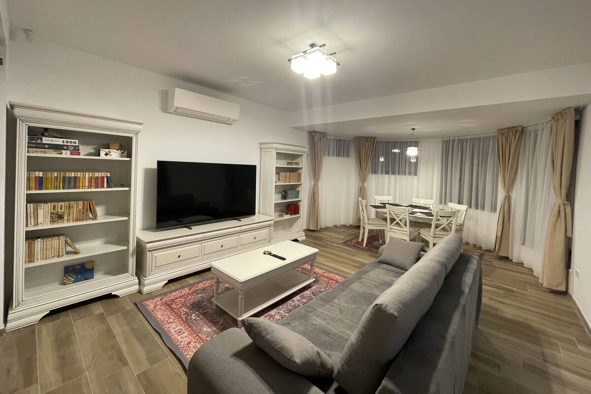 Sala de estar com televisão de tela plana, estante, sofá e área de jantar com mesa e cadeiras.