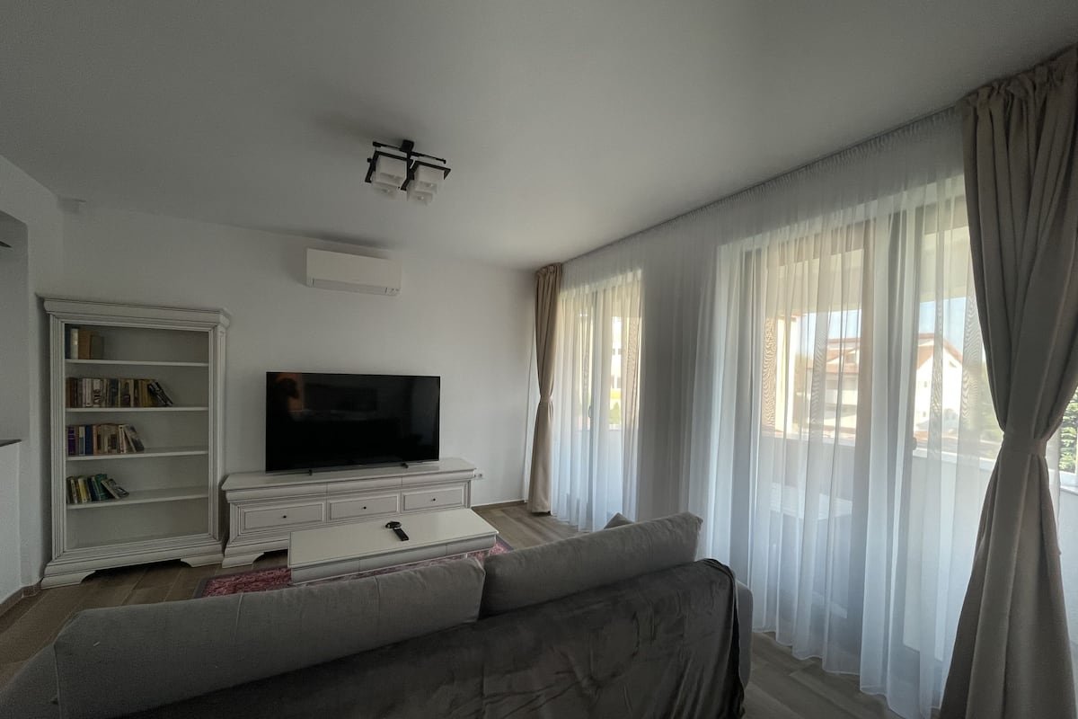 Uma sala de estar com uma televisão de tela plana sobre um móvel branco, um sofá cinza e uma estante de livros.