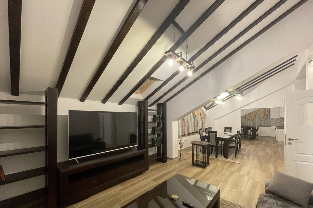 Uma sala de estar moderna com uma televisão de tela plana grande, móveis de madeira escura e uma área de jantar com mesa e cadeiras.