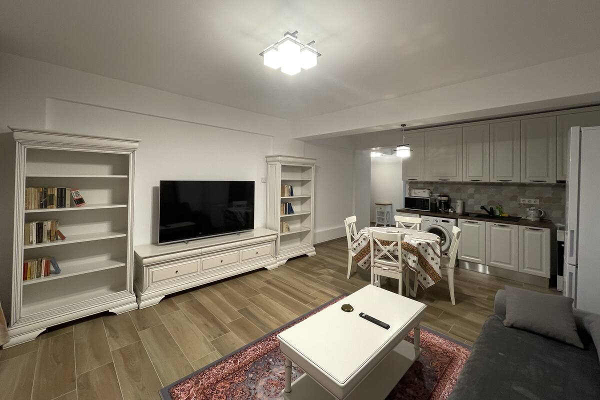 Uma sala de estar moderna com televisão de tela plana, estante, área de refeições com mesa e cadeiras, e cozinha com armários e eletrodomésticos.