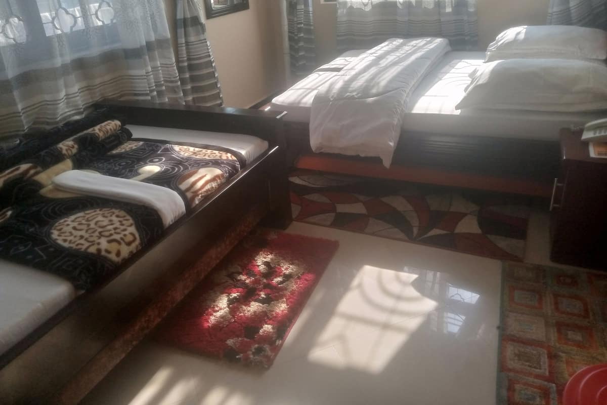 Quarto com duas camas, uma com colcha estampada e outra com colcha branca lisa. Há uma janela com cortinas, uma mesa pequena e um tapete estampado no chão.