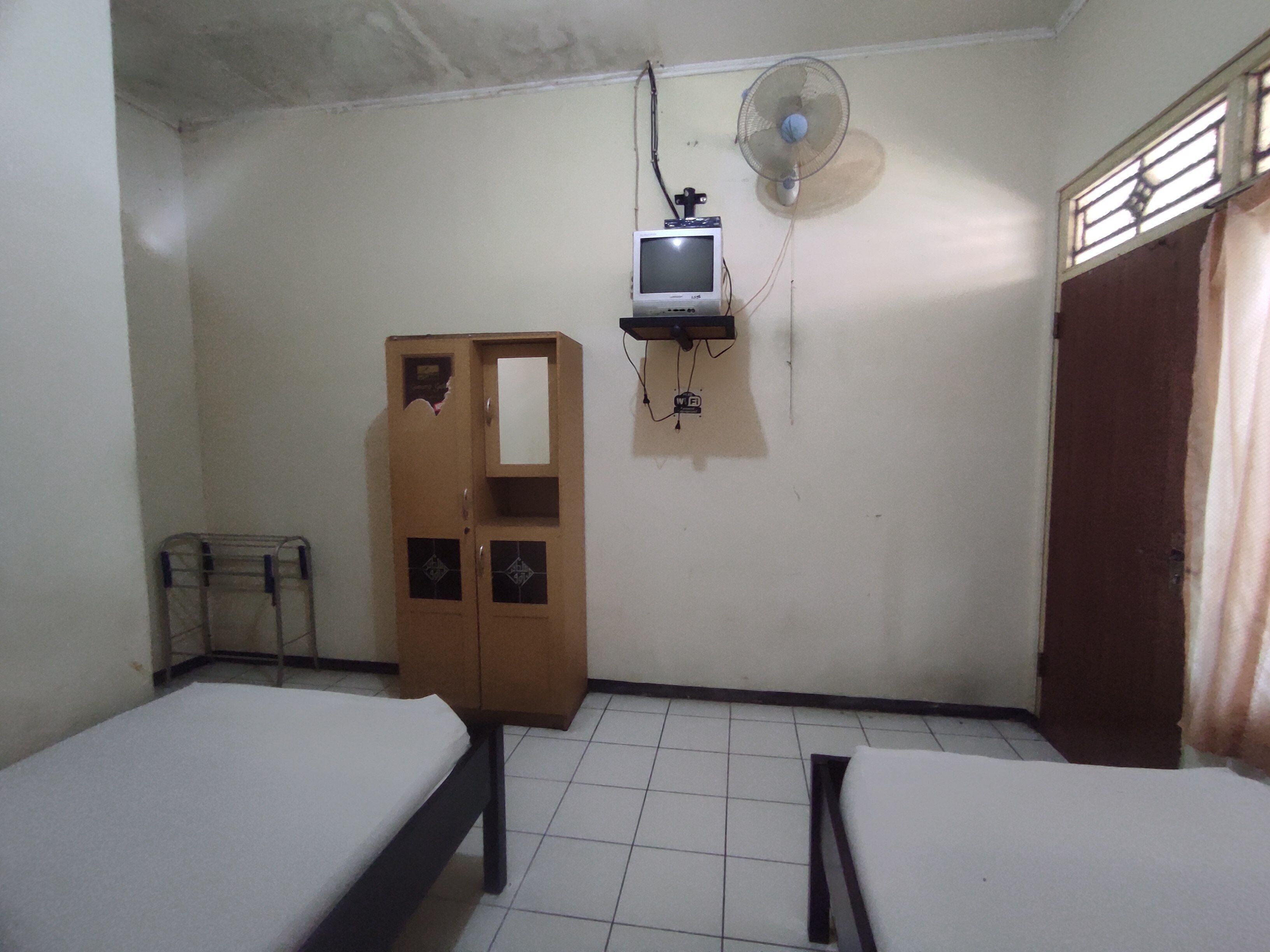 Um quarto pequeno e simples, com duas camas individuais, um guarda-roupa de madeira, uma mesa pequena, um ventilador fixado na parede e uma televisão.