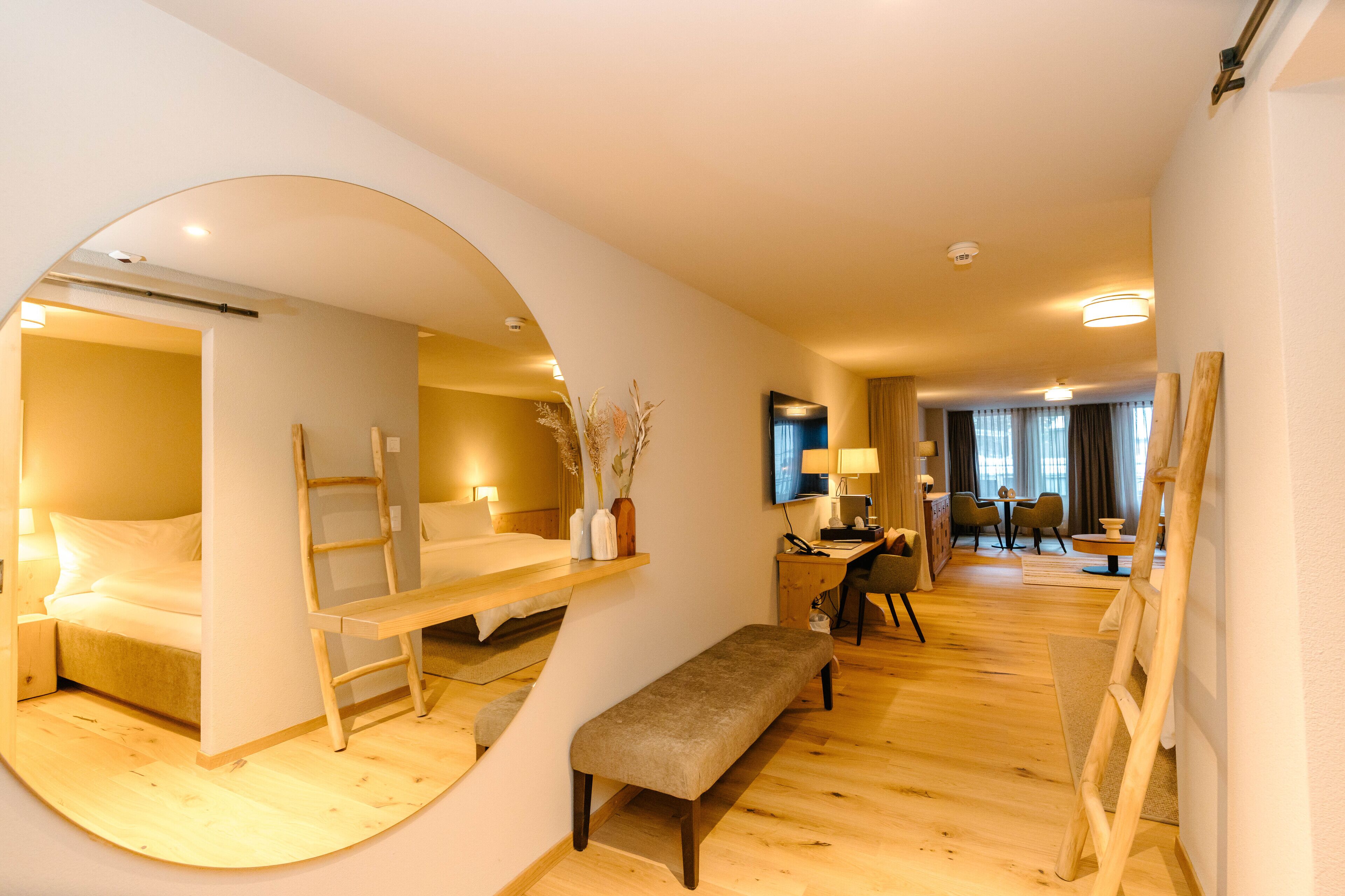 Quarto de hotel moderno com um espelho grande, uma cama, uma secretária com computador, um banco e um espaço de refeições com cadeiras e uma mesa.