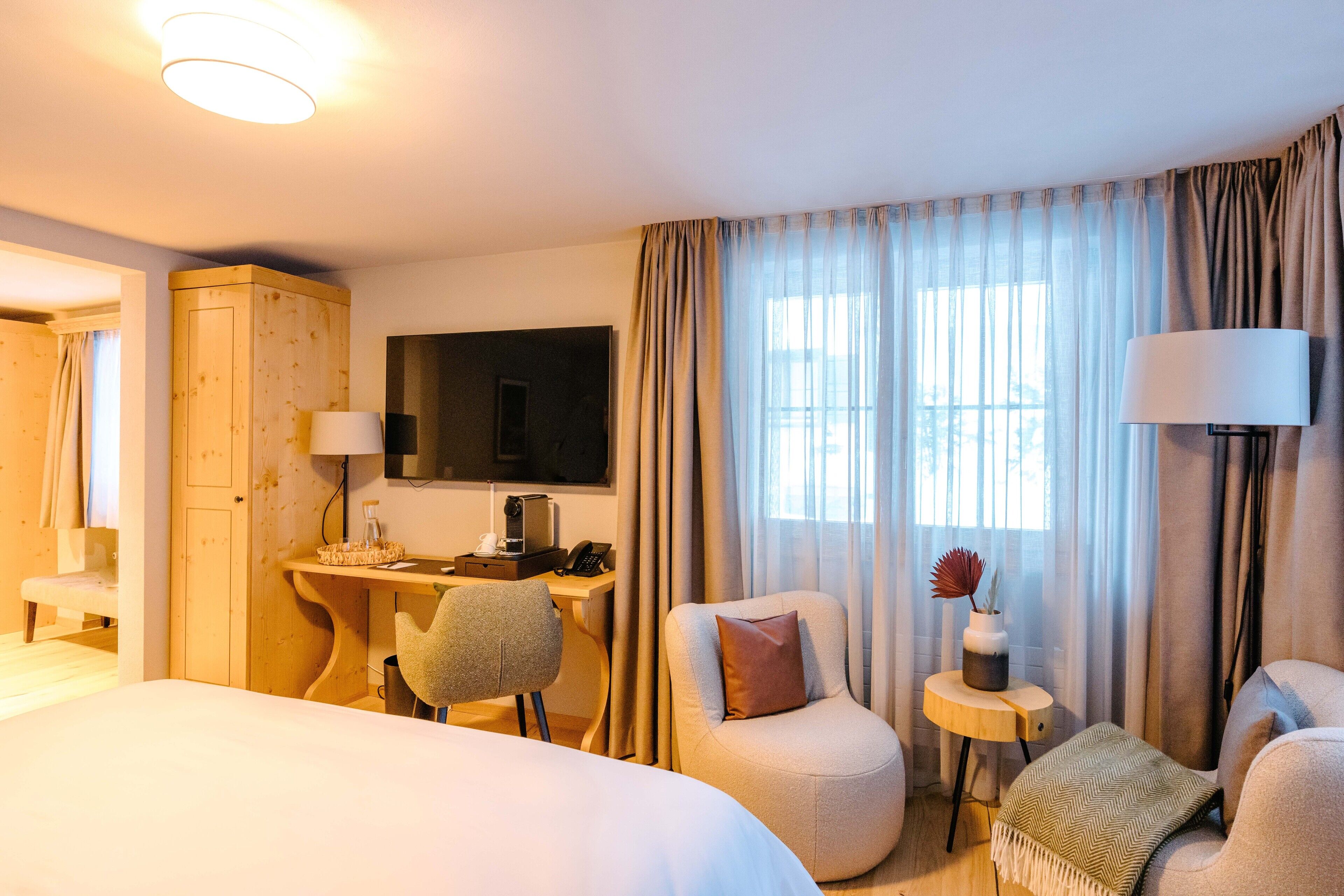 Quarto de hotel com cama, escrivaninha, cadeira, televisão e janela com cortinas.