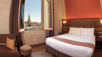 Quarto de hotel com uma cama grande, um sofá, um pequeno banquinho e uma janela com vista para um edifício.
