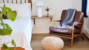 Um quarto com uma cama de madeira, uma poltrona de couro marrom, um banquinho redondo de fibra natural, uma mesa de cabeceira branca com uma luminária e um espelho na parede.