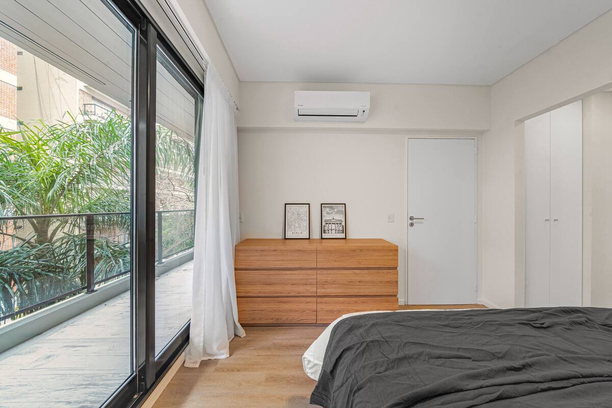 Quarto com uma janela grande, uma cama, uma cômoda de madeira e uma porta.