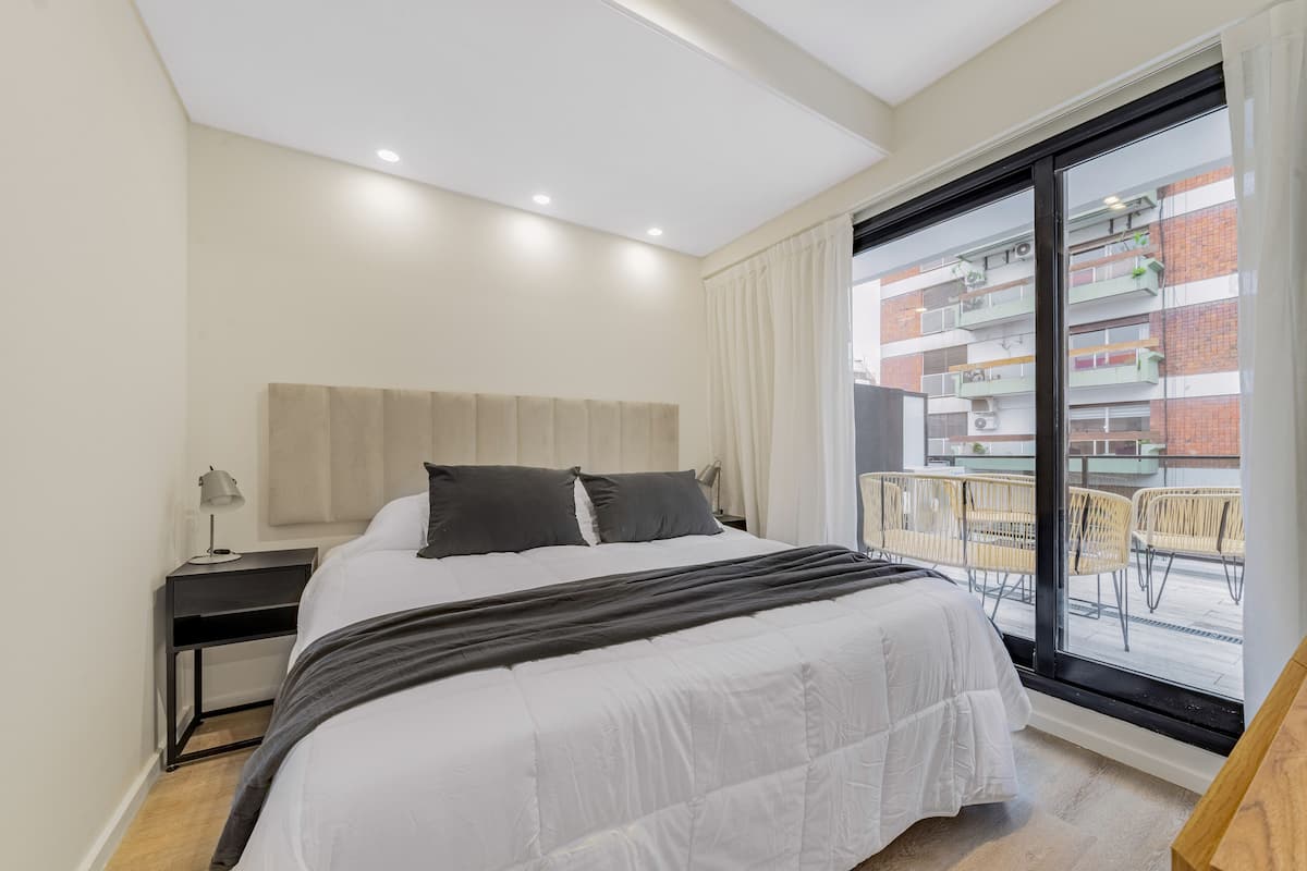 Quarto moderno com uma cama grande, uma mesinha de cabeceira e vista para uma varanda com móveis de exterior.