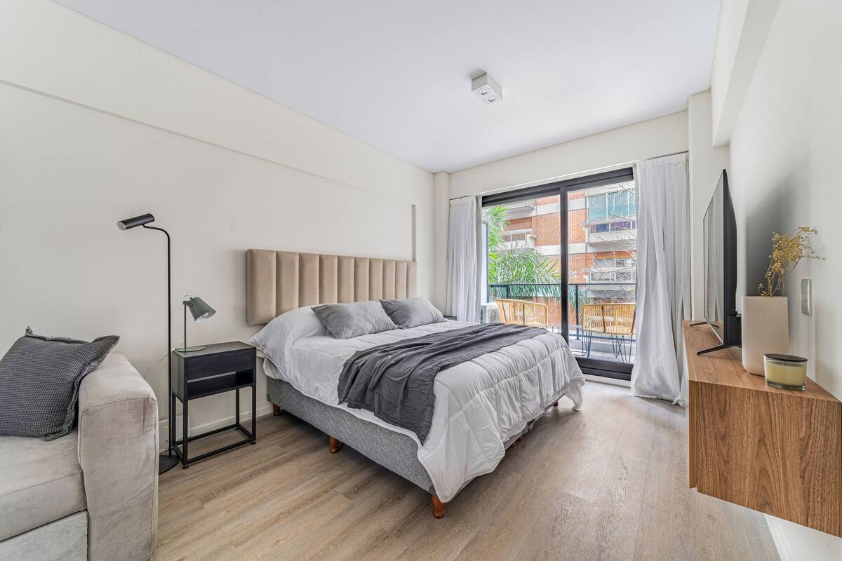 Quarto moderno com cama, mesa de cabeceira, luminária, cômoda de madeira e porta de vidro deslizante para uma varanda.