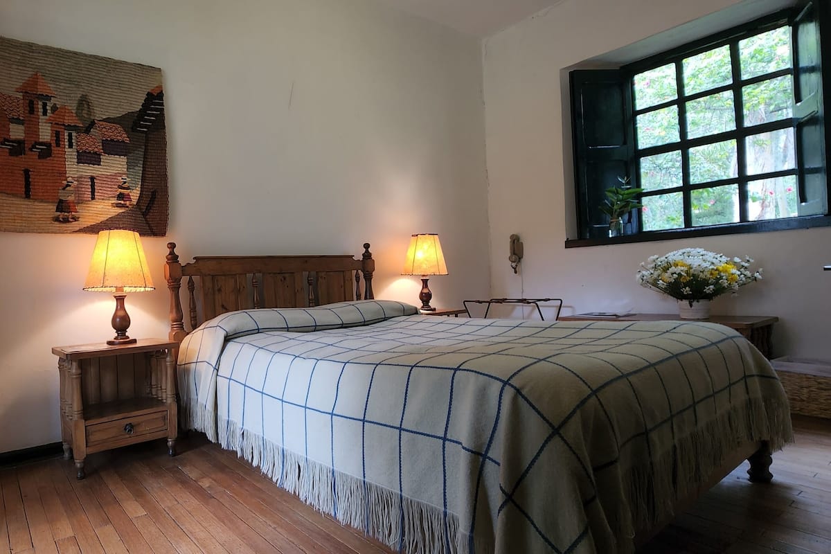 Um quarto com uma cama, duas mesas de cabeceira com luminárias, uma tapeçaria na parede e uma janela com vista para o verde.
