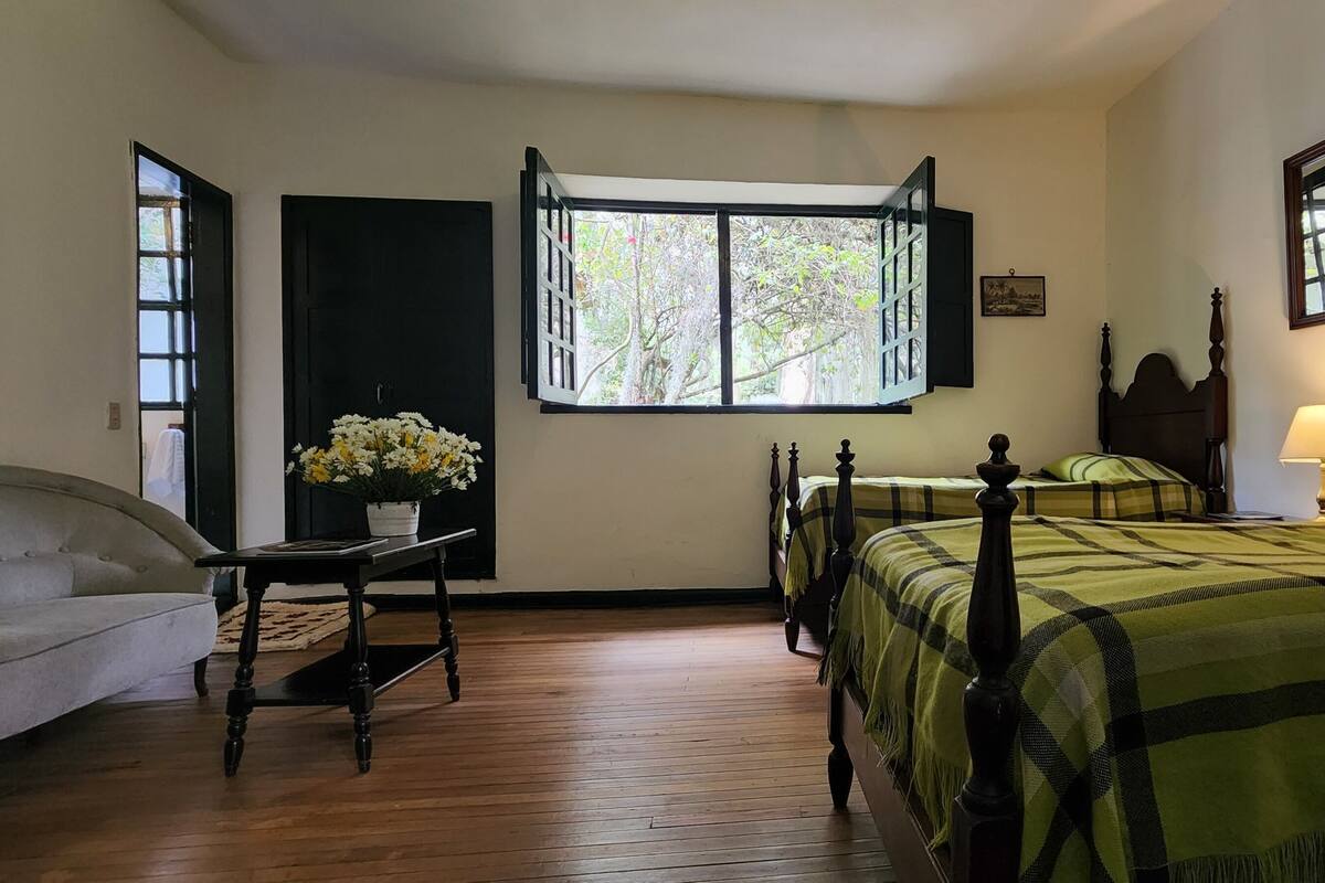 Quarto com duas camas, um sofá, uma janela com a persiana aberta, uma luminária e um espelho na parede.