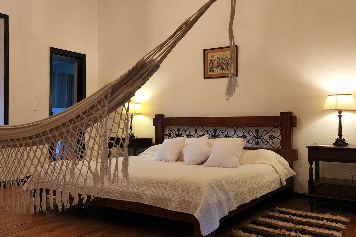 Quarto com rede, cama com roupa de cama branca, duas luminárias de cabeceira, mesa de cabeceira de madeira e uma moldura com um quadro na parede.
