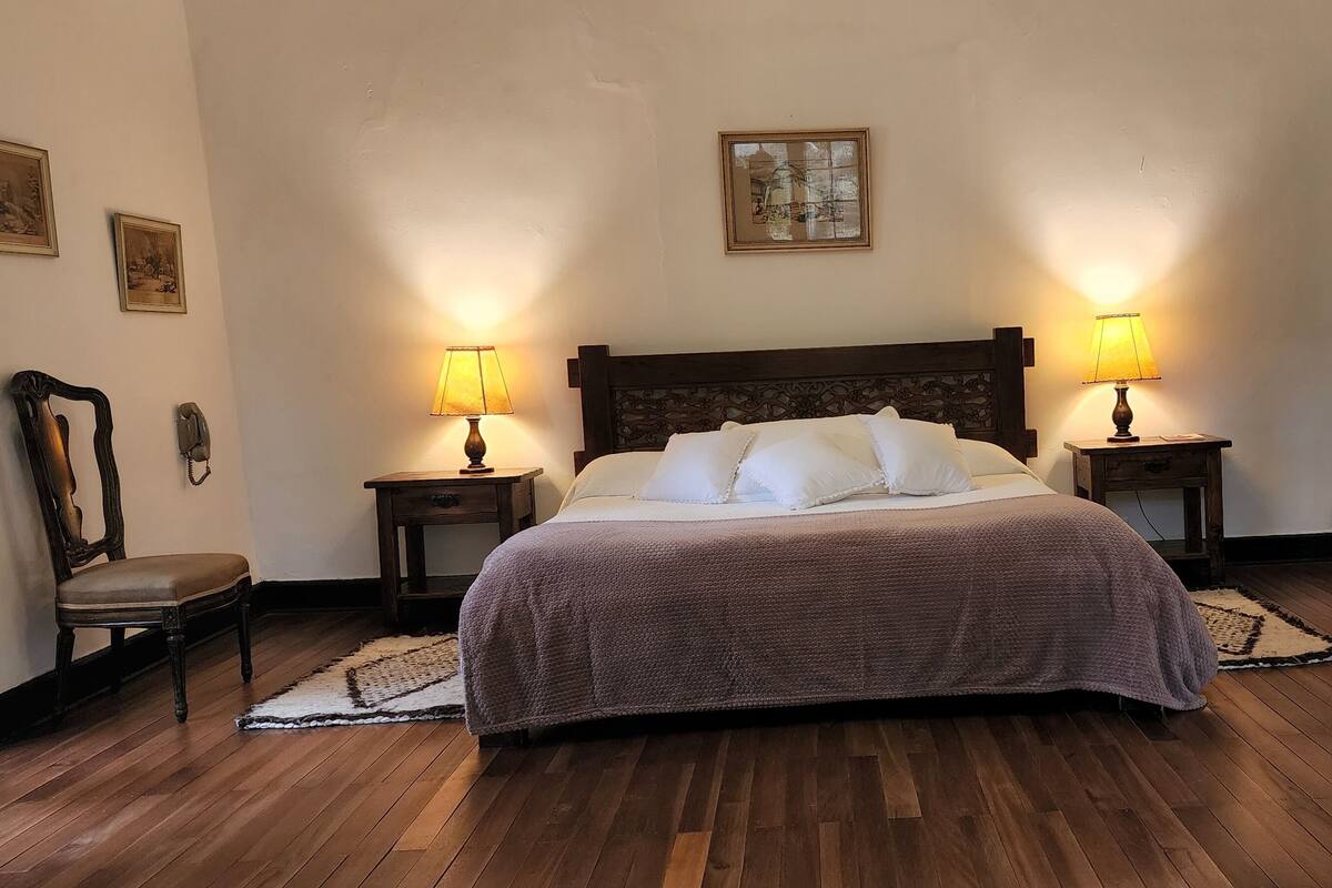 Um quarto bem organizado, com uma cama grande, duas mesas de cabeceira com abajures, uma cadeira de madeira e uma porta.