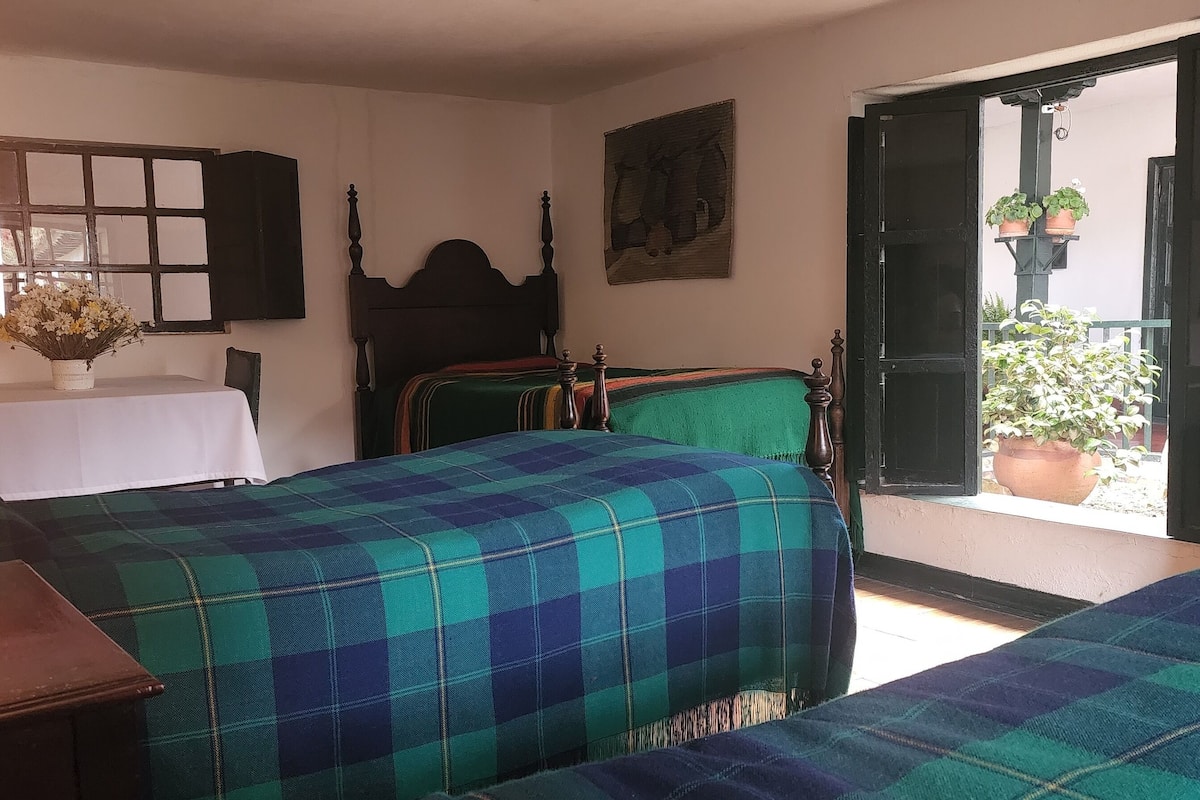 Quarto com duas camas, uma mesa com abajur, janela e porta que dá para área externa com plantas em vasos.