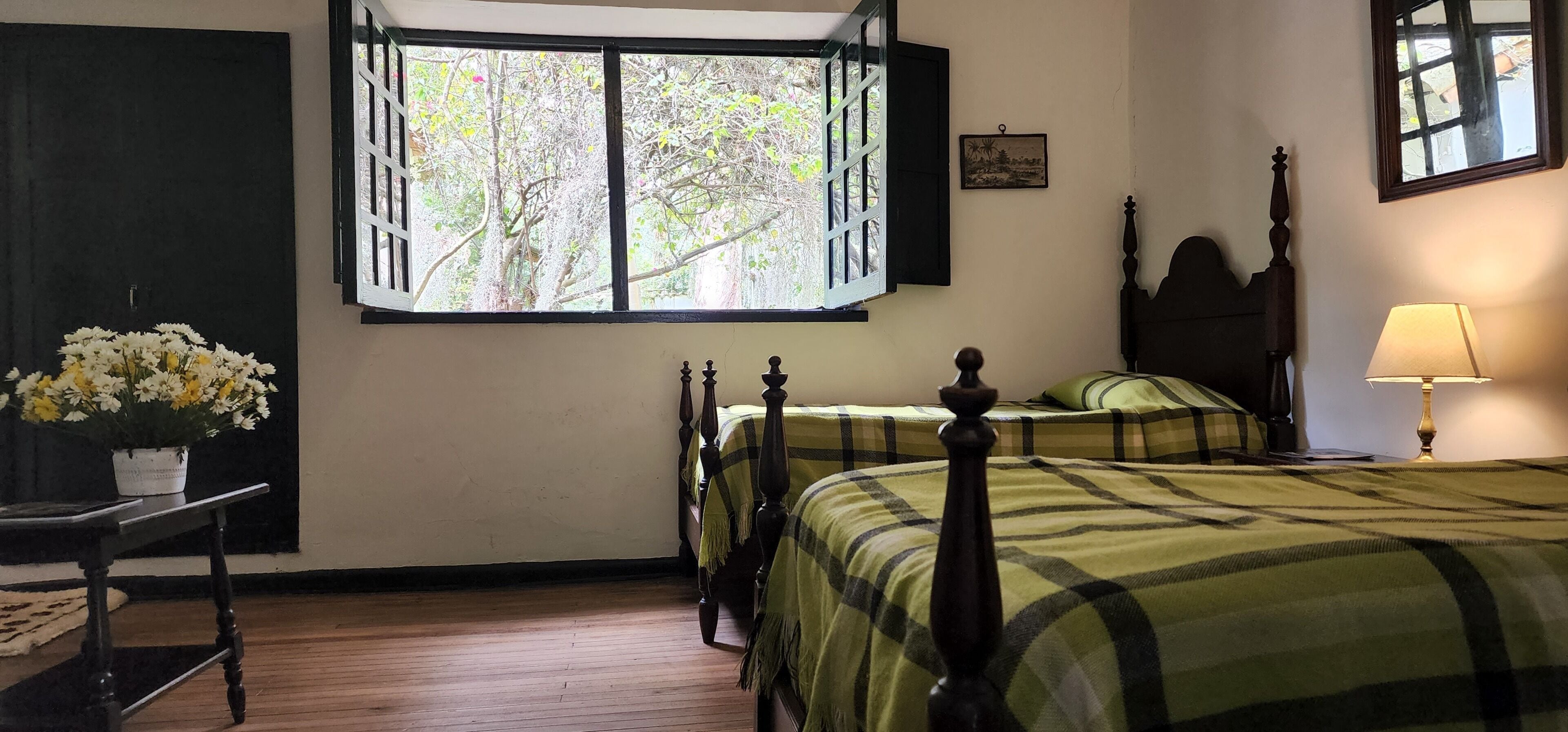 Quarto com duas camas, janela com vista para árvores, mesa de cabeceira com abajur e quadro na parede.