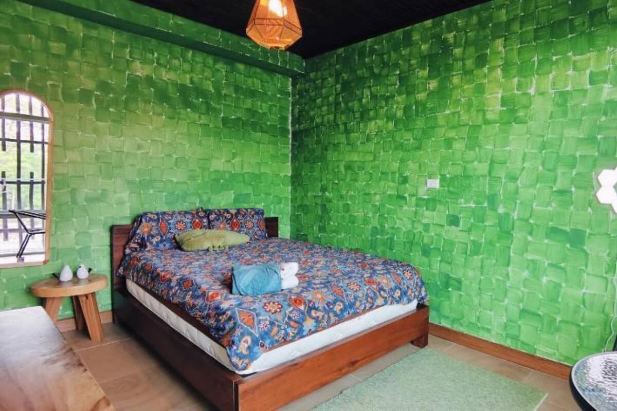 Quarto com parede em mosaico verde, cama de madeira com roupa de cama estampada, uma pequena mesa com um vaso de flores e um candeeiro pendente.