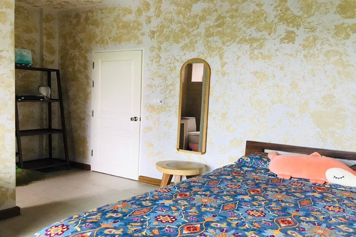 Um quarto com uma cama, um banquinho pequeno, uma estante e uma porta.
