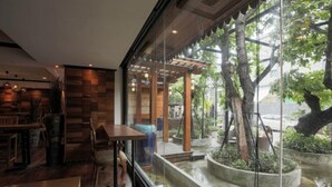 Uma área de refeições interna com mobiliário de madeira, divisória de vidro e vista para um jardim externo com um elemento aquático.