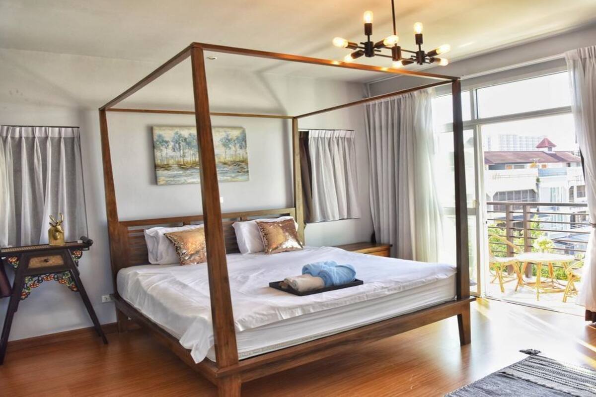 Uma cama de dossel com estrutura de madeira, roupa de cama branca e almofadas decorativas. Há um quadro na parede acima da cama, e uma pequena mesa com um objeto decorativo ao lado. O quarto possui uma varanda com vista para edifícios.