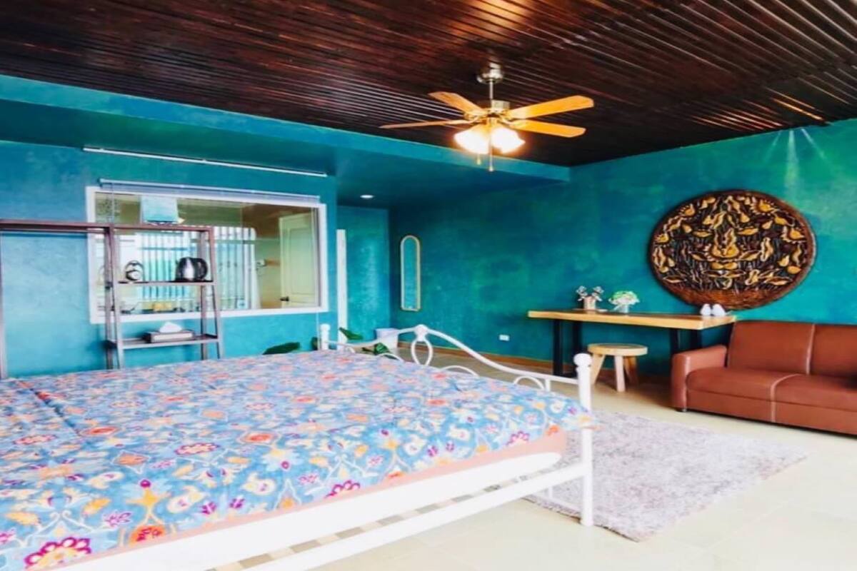 Quarto com cama, ventoinha no teto, mesinha, sofá e um objeto decorativo na parede.