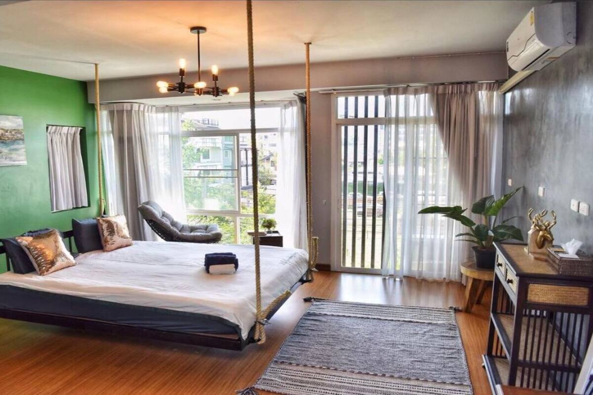 Quarto com uma cama grande, uma cadeira, uma escrivaninha com uma planta, e vista para o exterior através de grandes janelas.