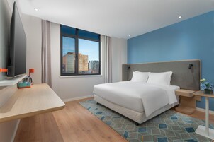 Quarto de hotel com uma cama, televisão, escrivaninha e janela com vista para a cidade.