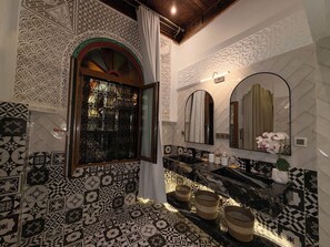Casa de banho com azulejos estampados, uma janela grande com moldura decorativa, bancada de mármore e espelho com moldura ornamental.