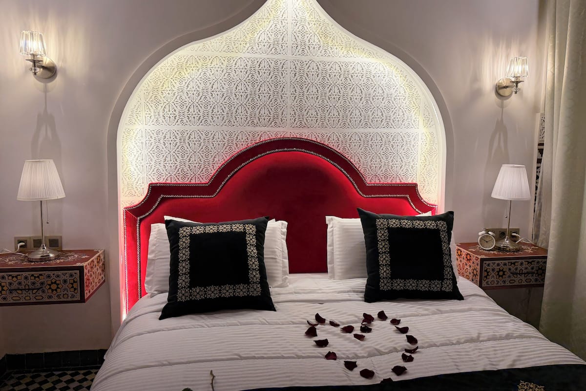 Quarto de hotel com cabeceira estofada em vermelho, parede branca com padrão e duas mesas de cabeceira com luminárias.