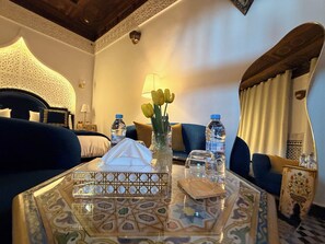 Quarto de hotel com uma cama, dois sofás, uma mesa de centro com bandeja decorativa, um vaso com flores amarelas e garrafas de água.