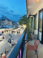Uma varanda com vista para um mercado de rua movimentado, um edifício moderno com grandes janelas e um edifício tradicional ao fundo.