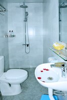 Casa de banho com sanita, lavatório com pétalas de rosa, chuveiro e prateleira de vidro com artigos de higiene pessoal.