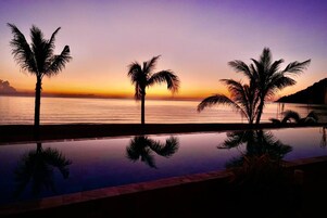 Uma piscina refletindo palmeiras e um pôr do sol sobre o oceano.