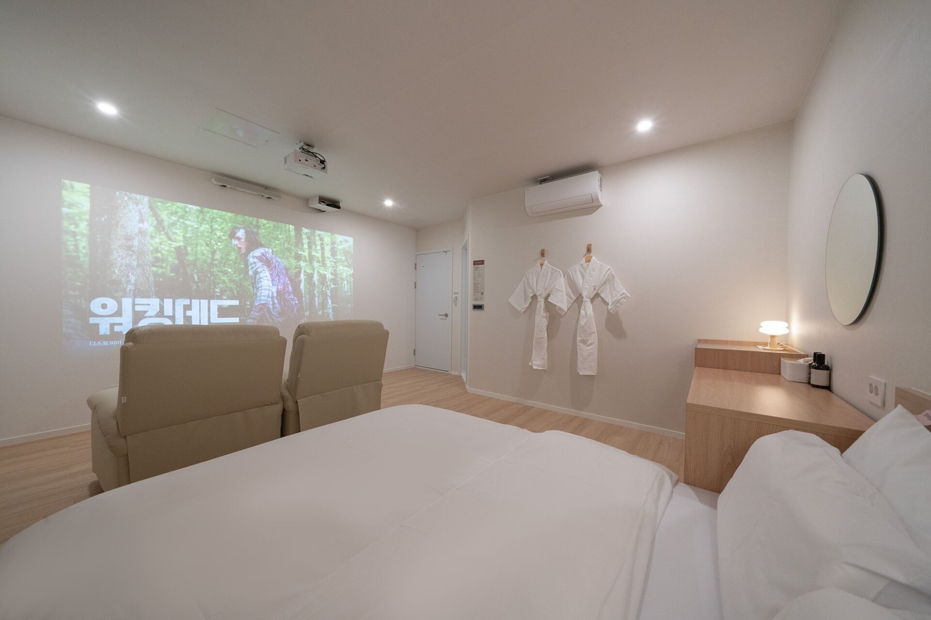 Quarto de hotel com cama, duas cadeiras, uma mesinha de centro com abajur, espelho e ar-condicionado fixado na parede.