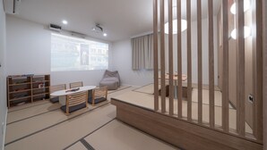 Quarto com tatames, uma mesa de madeira baixa, um sofá, tela de projetor e uma porta deslizante.