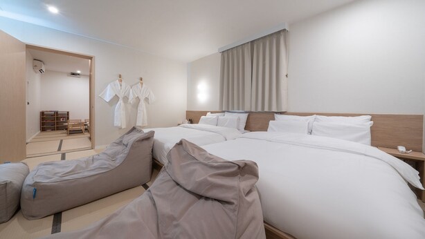 Quarto de hotel moderno com uma cama grande, uma mesinha de cabeceira, poltronas e um espaço de estar com almofadas.
