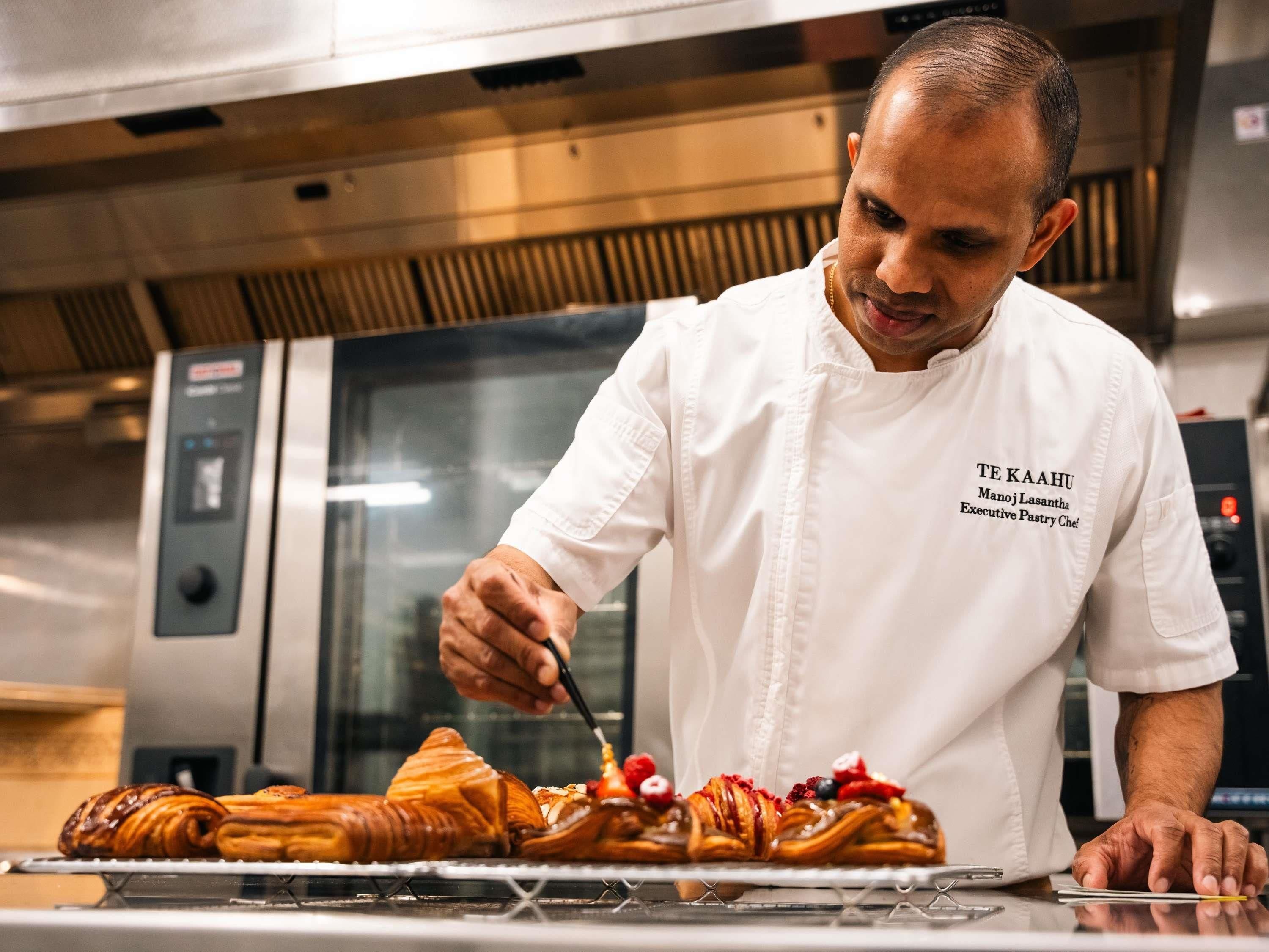 Um chef com uniforme branco, com 'TE KAAHU' e 'Manoj Lasantha Executive Pastry Chef' no peito, prepara doces em uma cozinha industrial equipada com eletrodomésticos de aço inoxidável e uma janela de vidro.