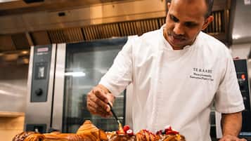 Um chef com uniforme branco, com 'TE KAAHU' e 'Manoj Lasantha Executive Pastry Chef' no peito, prepara doces em uma cozinha industrial equipada com eletrodomésticos de aço inoxidável e uma janela de vidro.