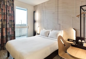 Quarto de hotel com uma cama grande, uma escrivaninha, uma cadeira, um abajur e vista para o exterior.