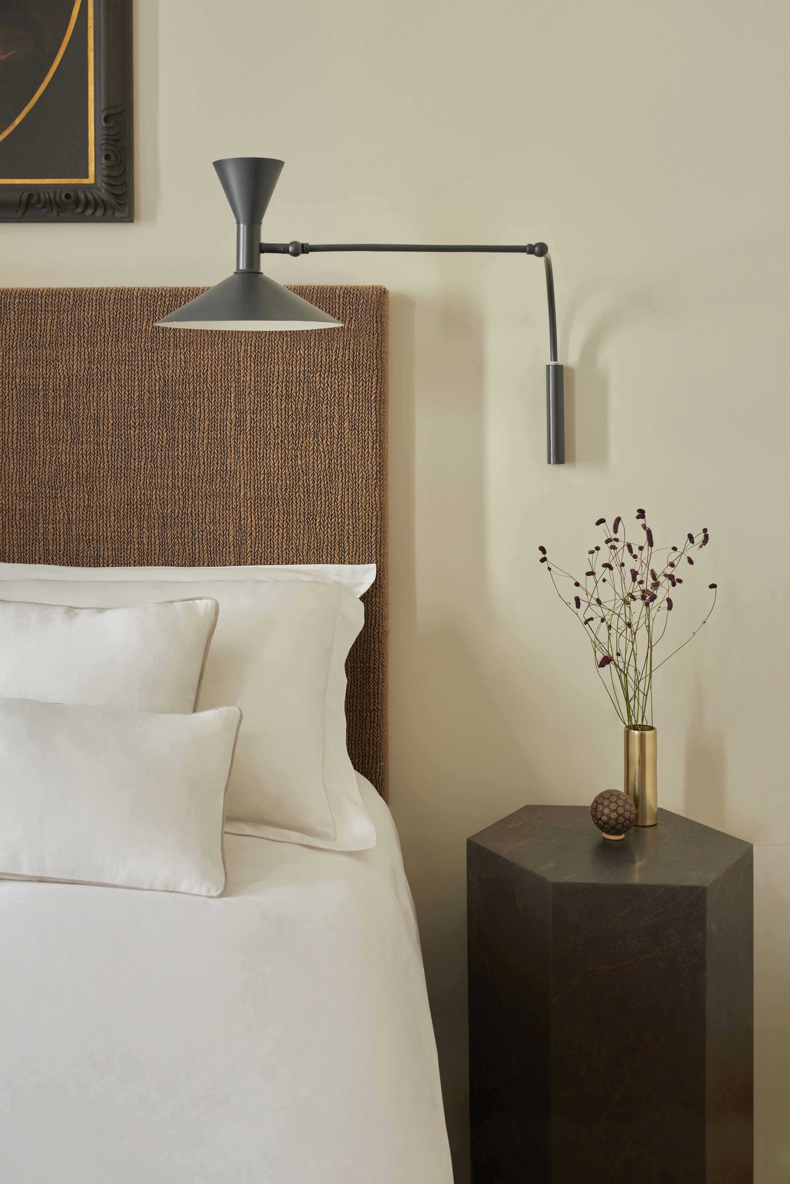 Uma cama bem arrumada com lençóis e almofadas brancas, uma luminária de chão moderna e um pequeno vaso com flores secas sobre uma mesinha lateral escura.