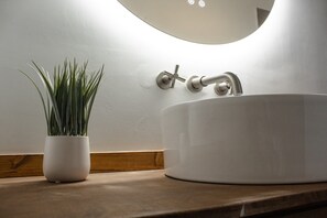 Uma pia branca com uma torneira moderna, uma planta em vaso e uma bancada de madeira em frente a uma parede lisa.