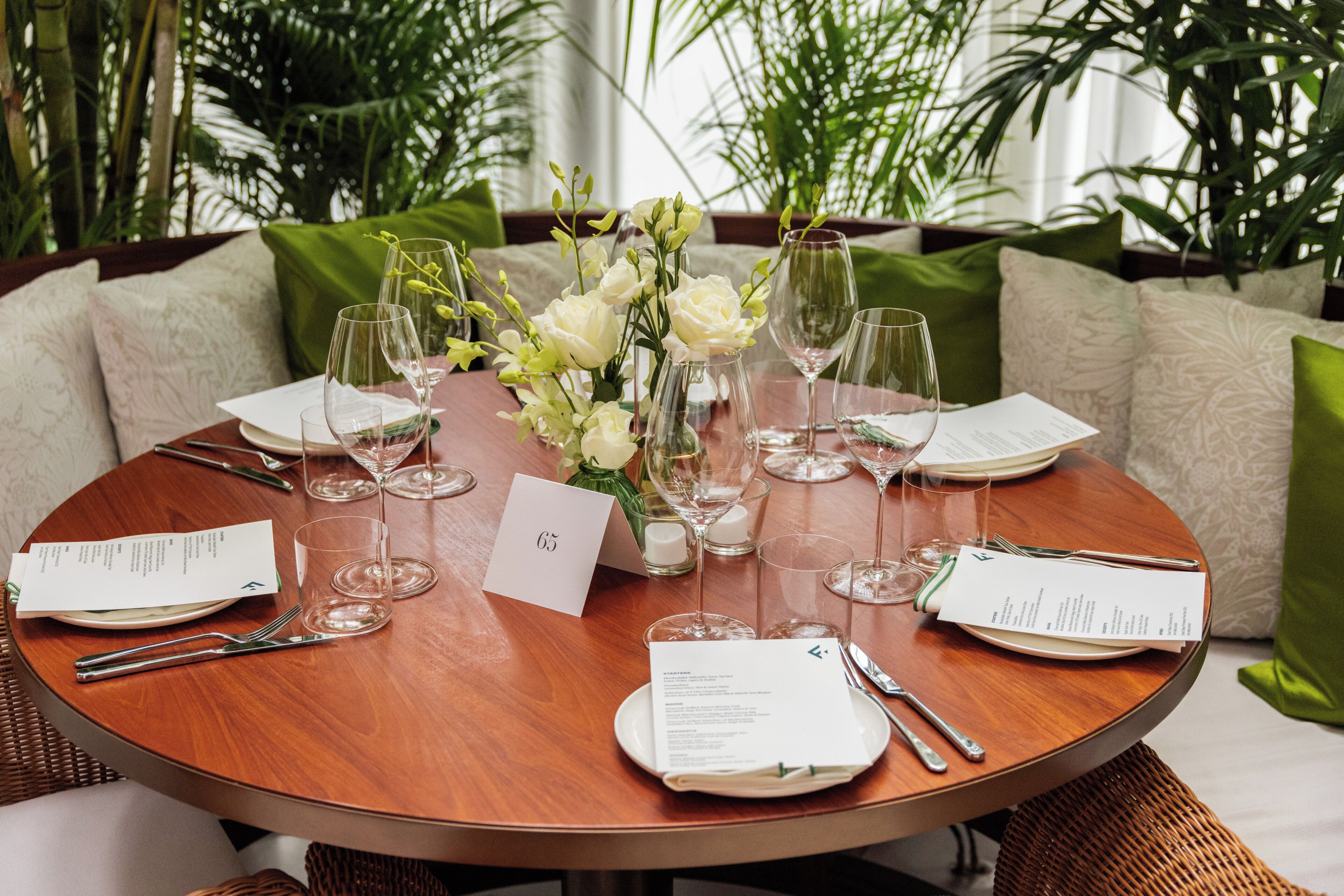 Mesa de jantar redonda posta com pratos brancos, copos de vinho transparentes e um arranjo de flores brancas.