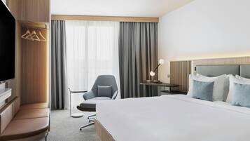 Quarto de hotel com cama, área de estar com uma cadeira, TV, escrivaninha com abajur e janela com cortinas.