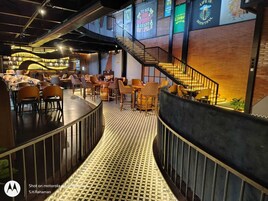 Um restaurante moderno com área de bar central, diversas opções de assento e escada que leva a um andar superior.