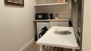 Cozinha compacta com micro-ondas, cafeteira e uma pequena geladeira. Há uma bancada branca com uma tigela e uma cadeira. Prateleiras acima contêm caixas e um armário pequeno.